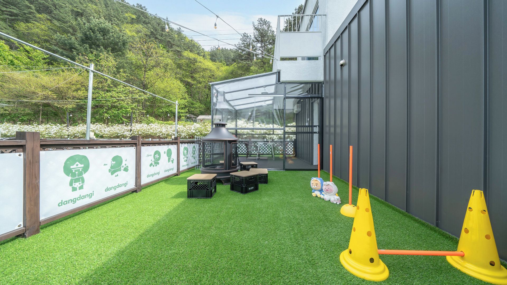 Área externa com grama sintética, estrutura de vidro e parquinho infantil com cones amarelos.