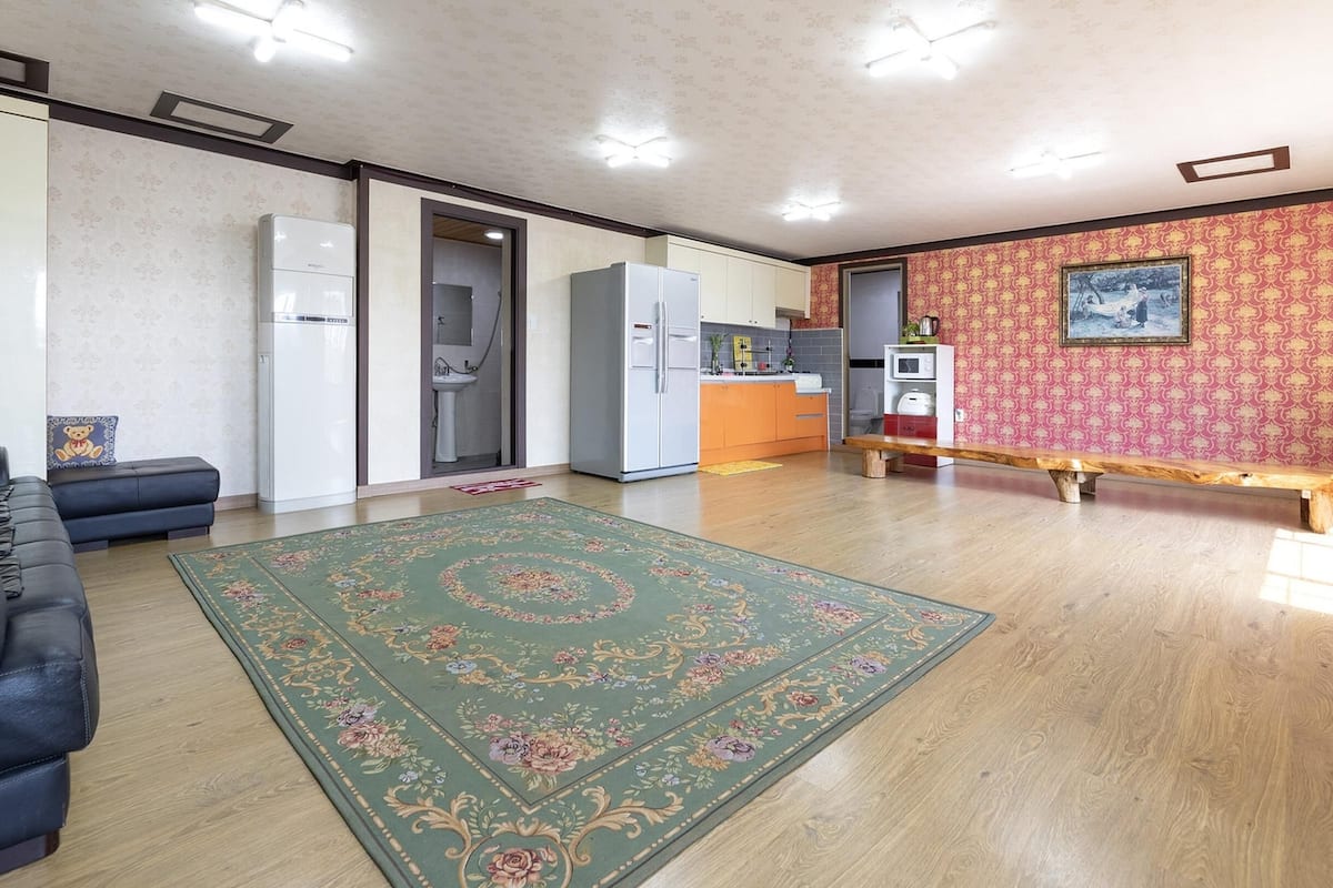 Uma sala de estar ampla com um sofá, um tapete floral verde, um banco de madeira, uma cozinha com geladeira e uma parede estampada.