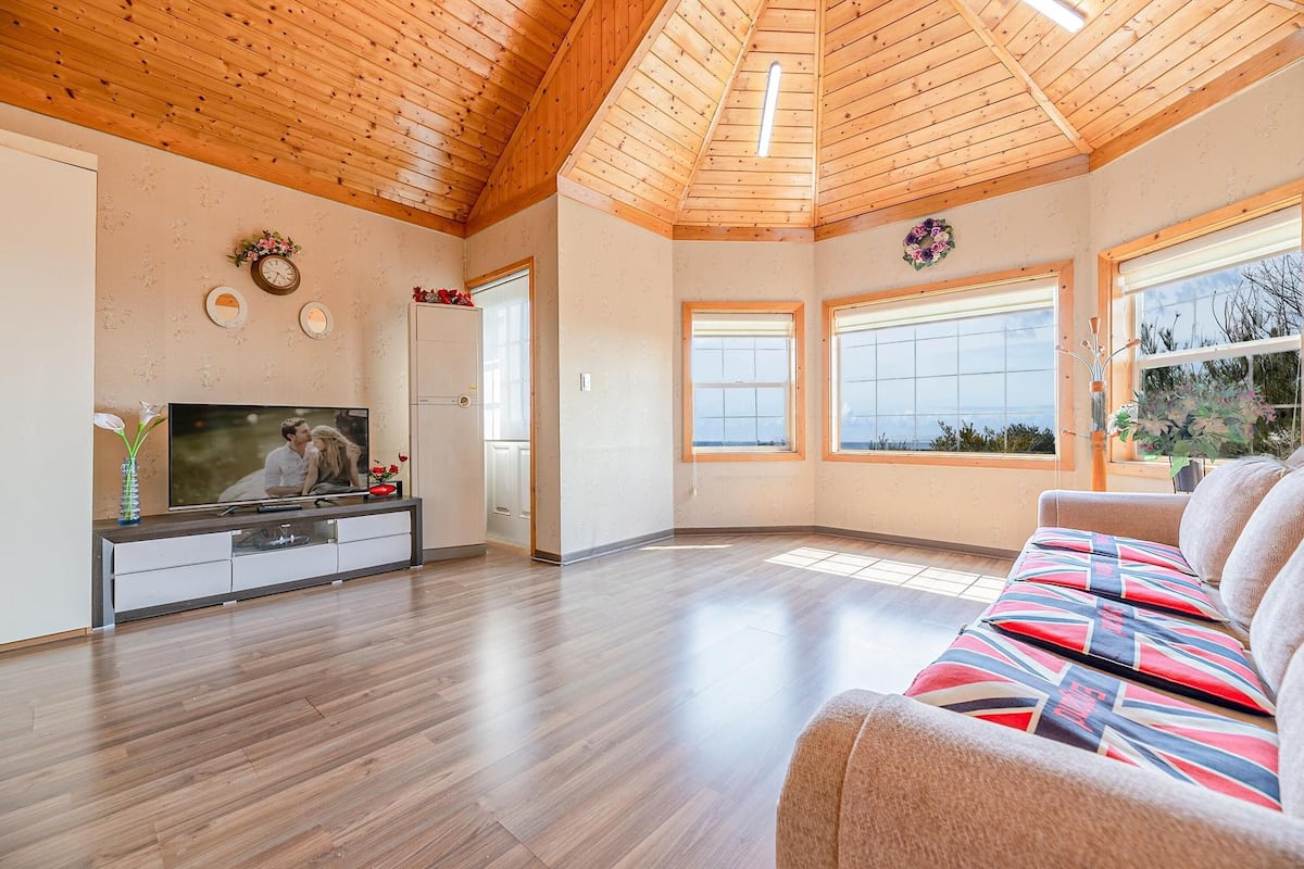 Uma sala de estar espaçosa com teto de madeira, janelas amplas, TV de tela plana e sofá com almofadas estampadas.