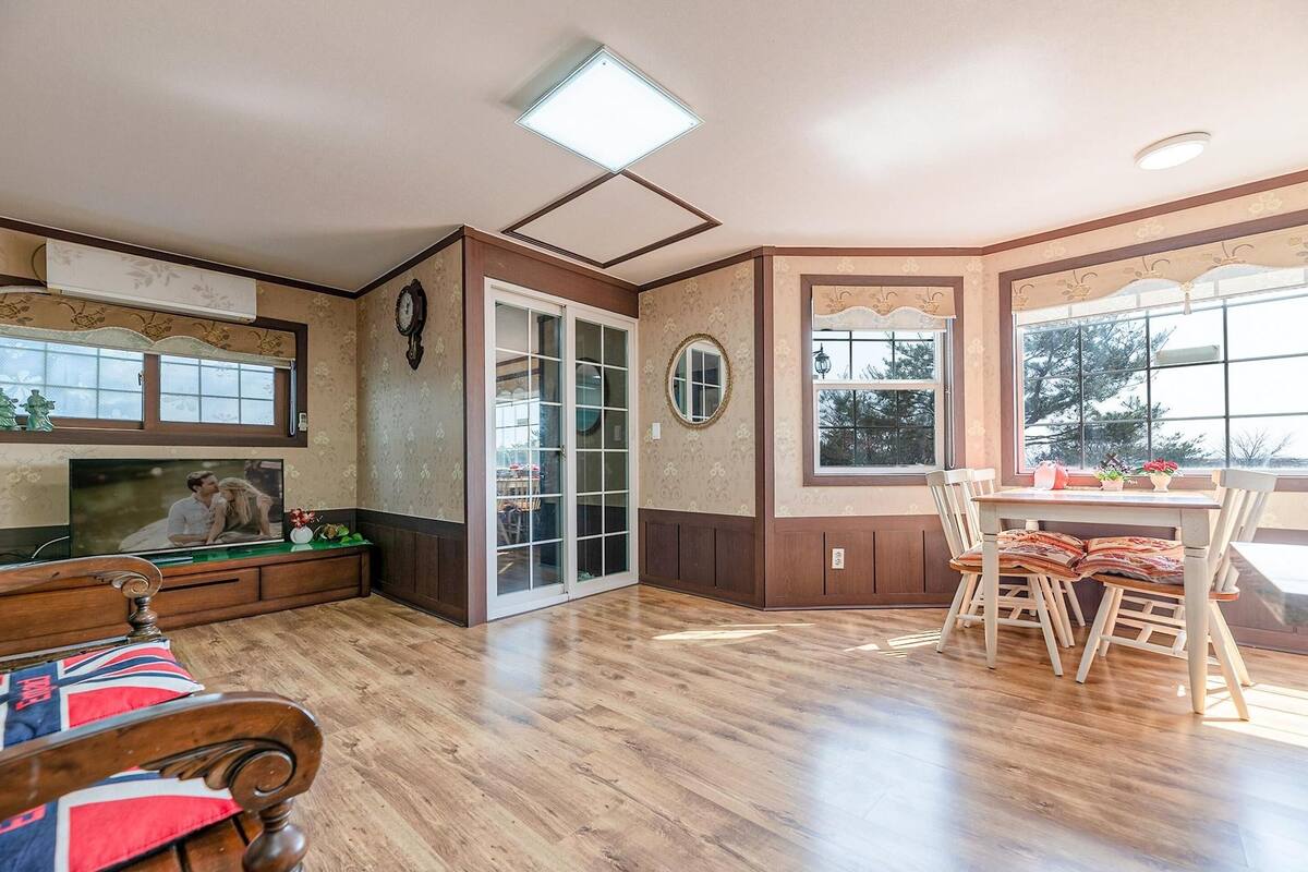 Uma sala de estar com piso de madeira, uma televisão, uma mesa de jantar com cadeiras e uma janela grande.