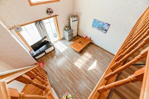Quarto com piso de madeira, uma pequena mesa, geladeira, janela com cortinas e uma escada.