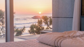 Quarto com vista para o oceano ao pôr do sol, uma cama com travesseiro e janela com vista para o mar e pedras.
