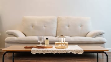 Uma mesa de madeira com toalha de renda, um copo, uma garrafa e um candeeiro de madeira sobre um soalho de madeira, com um sofá bege ao fundo.