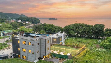 Um edifício moderno com piscina no terraço, cercado por vegetação exuberante e vista para o mar.