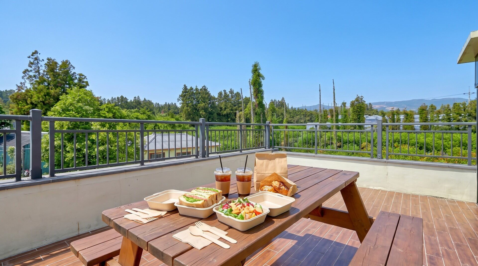 Um deck de madeira com mesas de piquenique, recipientes de comida e bebidas, com vista para uma paisagem pitoresca com árvores e céu claro.