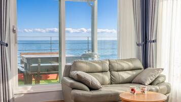 Quarto com sofá de couro, uma mesa com duas canecas e vista para o mar através de uma grande janela.