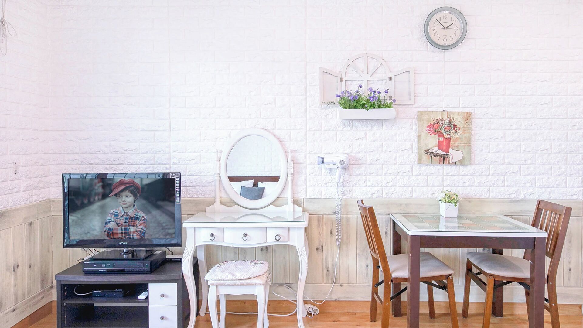 Quarto com televisão, penteadeira com espelho, relógio e área de refeições com cadeiras e mesa.