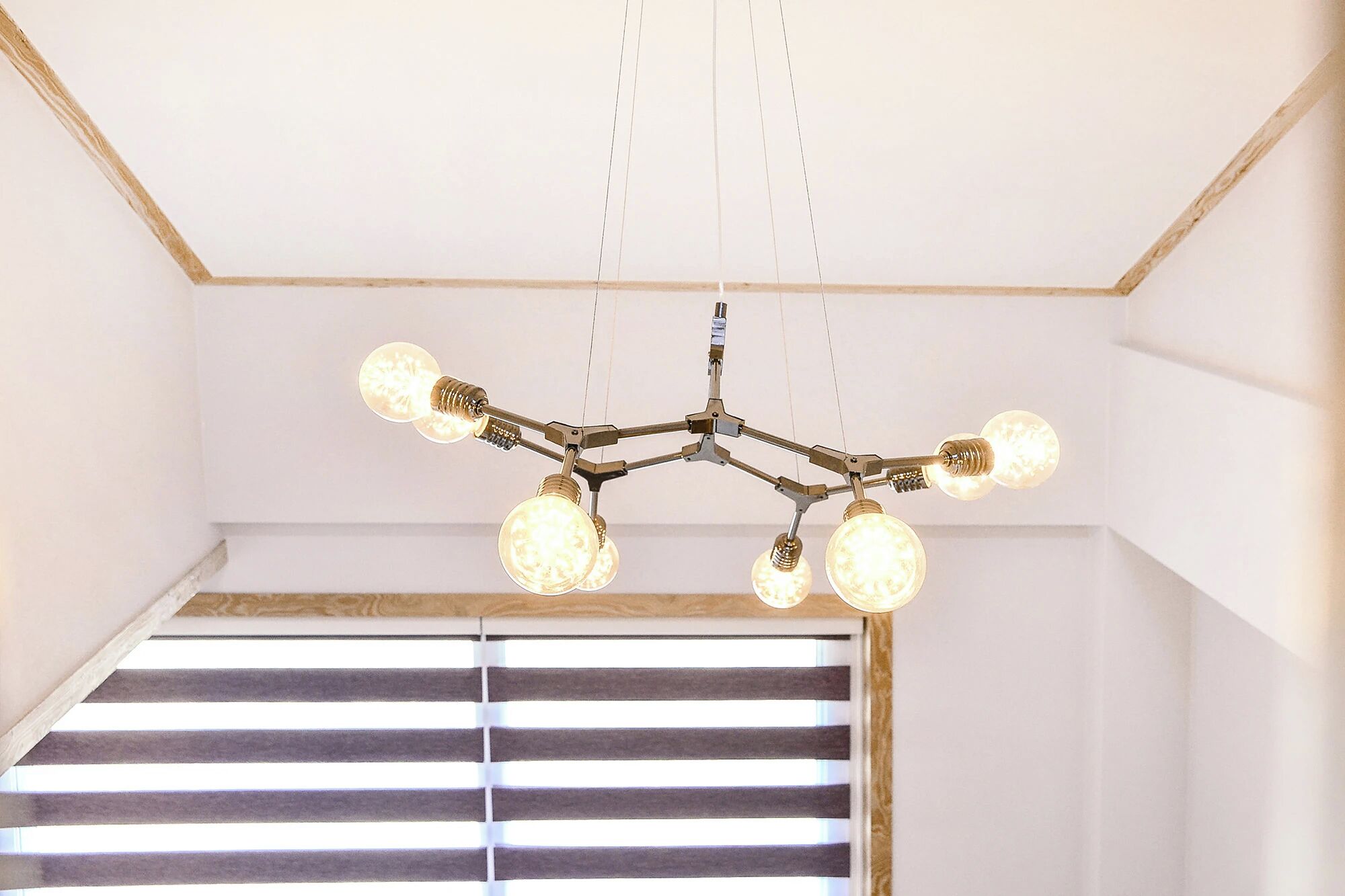 Um candelabro com várias lâmpadas em um quarto com paredes brancas e uma janela com venezianas horizontais.