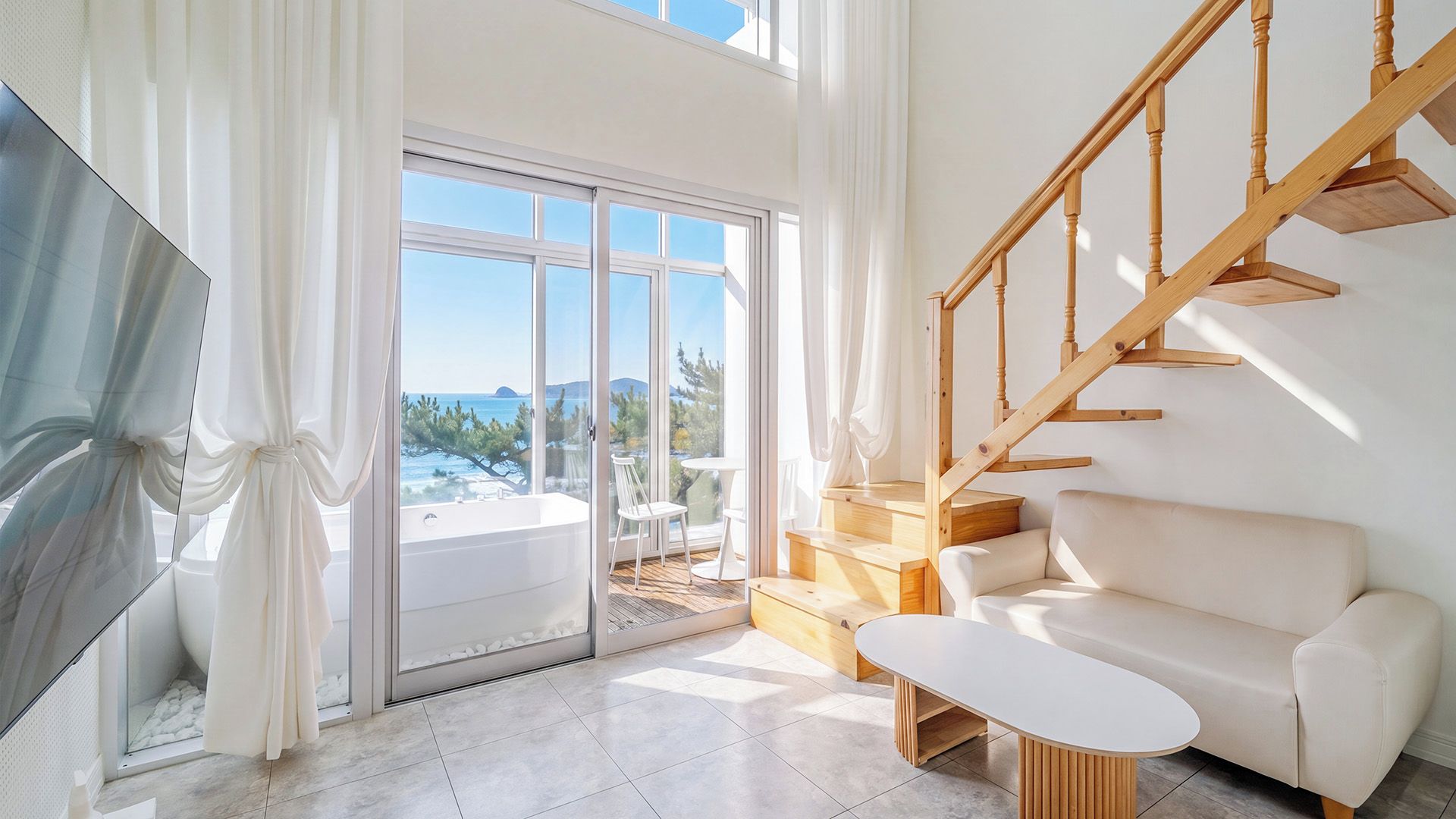 Ampla sala de estar com escada de madeira, sofá branco e vista para o mar através de grandes janelas.