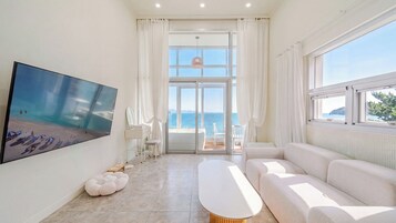 Sala de estar moderna com uma TV grande, sofá, mesa de centro e vista para o mar através de uma porta de vidro.