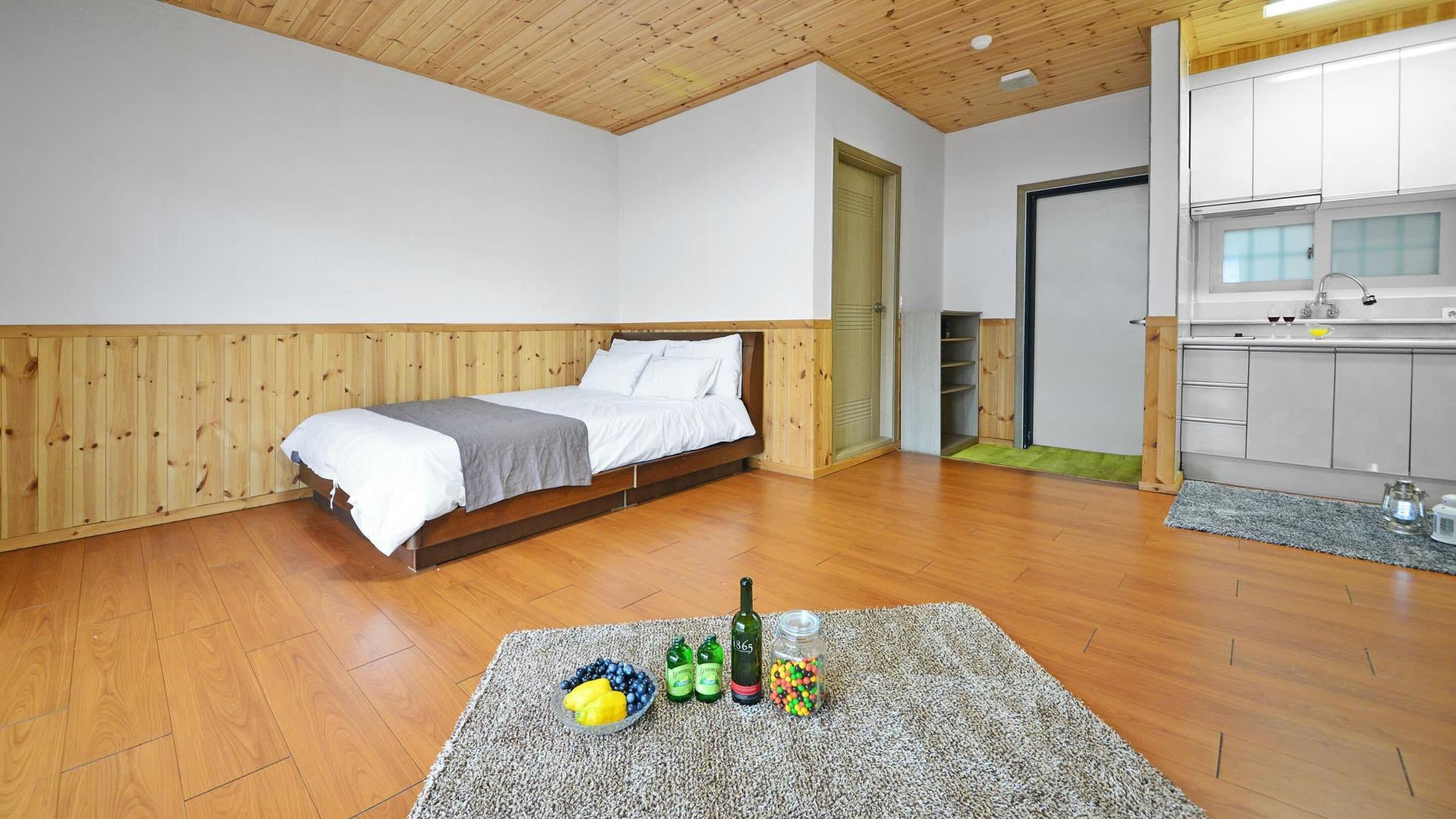 Quarto com cabeceira de madeira, cama com roupa de cama branca, pequena área de cozinha com armários brancos e tapete com garrafas e lanches no chão.