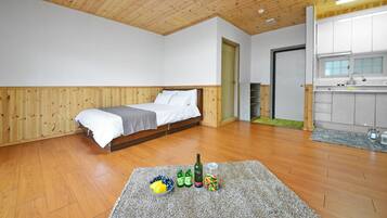 Quarto com cabeceira de madeira, cama com roupa de cama branca, pequena área de cozinha com armários brancos e tapete com garrafas e lanches no chão.