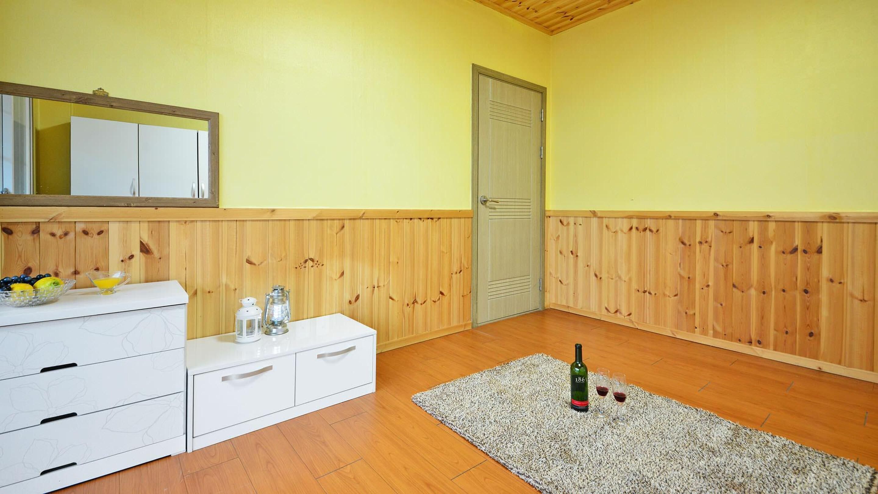 Quarto com painéis de madeira, penteadeira branca, espelho, porta e tapete com uma garrafa e copos.