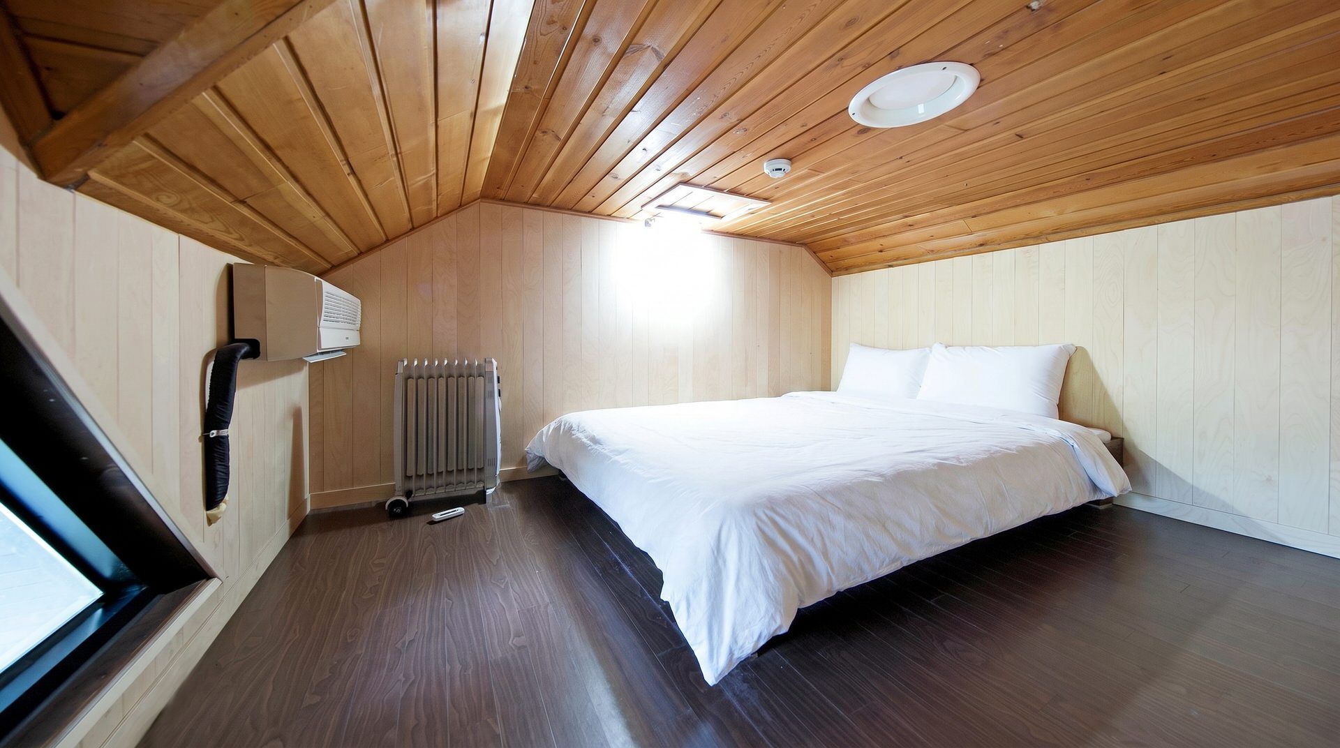 Quarto com teto de madeira, radiador, cama com roupa de cama branca e janela.