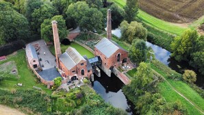 Vista aérea de um edifício industrial histórico com duas chaminés, um canal e vegetação ao redor.