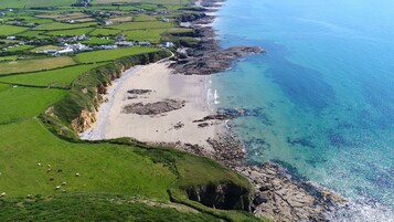 Vista aérea de uma área costeira com praia arenosa, margem rochosa e campos verdes.