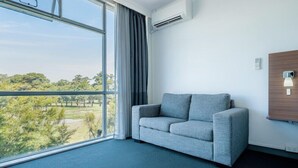 Quarto de hotel moderno com um sofá cinzento, uma janela grande com cortinas e vista para um campo de golfe.
