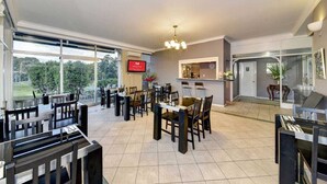 Uma ampla sala de jantar com várias mesas e cadeiras, grandes janelas com vista para o exterior, e uma cozinha moderna ao fundo.