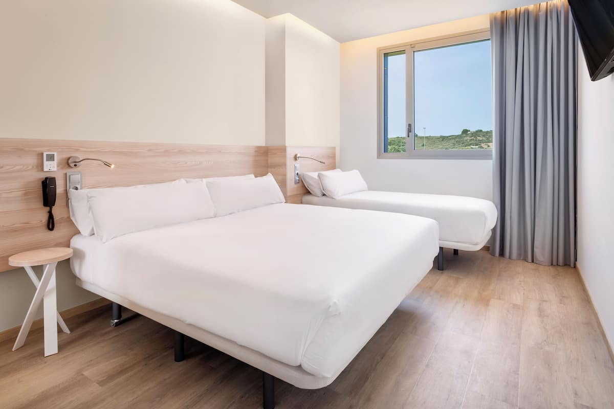 Quarto de hotel com uma cama grande, uma cama menor, uma mesa de cabeceira, um telefone e uma janela com vista para o exterior.