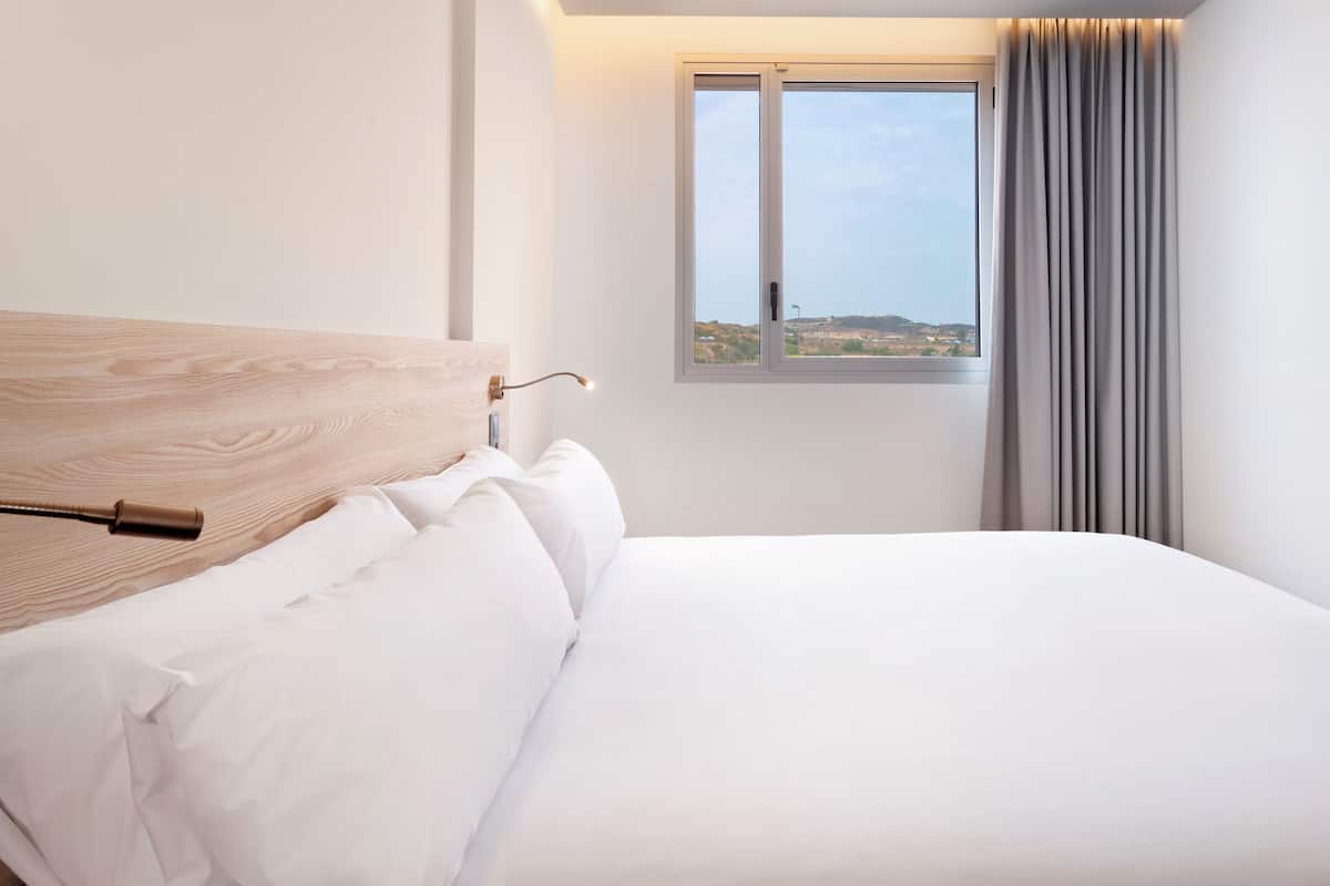 Cama bem arrumada com lençóis brancos e cabeceira de madeira em quarto de hotel, com janela grande e cortinas.
