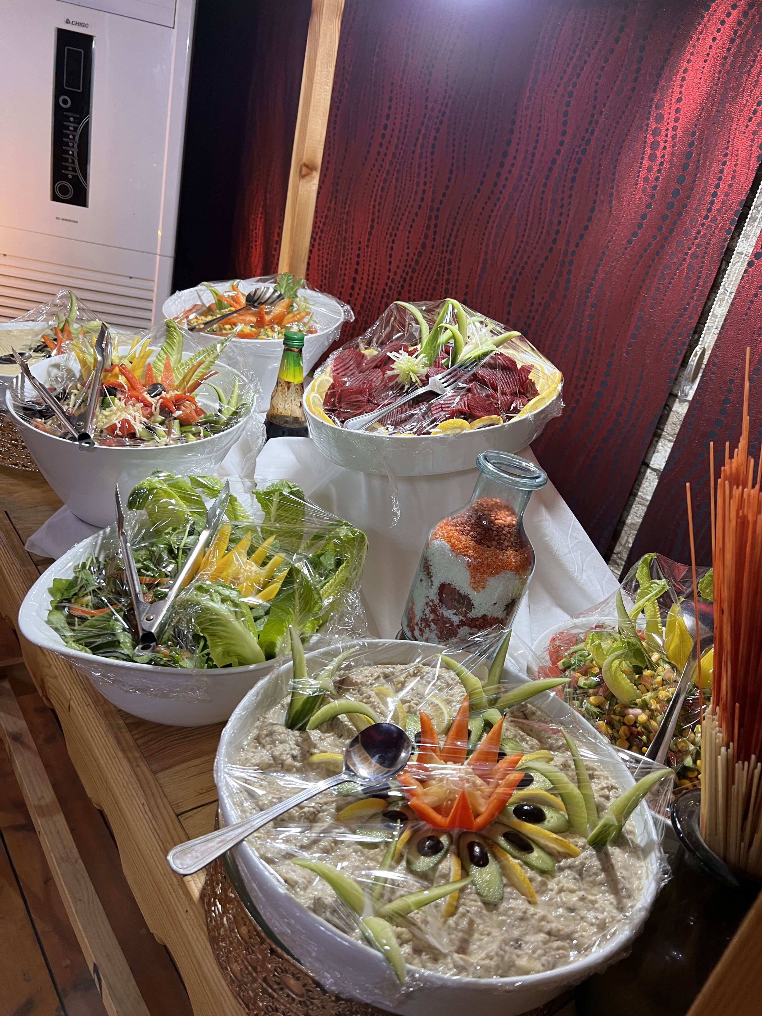 Uma mesa de buffet com diversos pratos, incluindo saladas, legumes e um prato de carne, coberta com filme plástico.