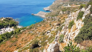 Paisagem costeira com terreno rochoso, mar azul cristalino e alguns edifícios ao longe.