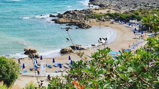 Uma praia com guarda-sóis e espreguiçadeiras, costa rochosa e vegetação ao primeiro plano.