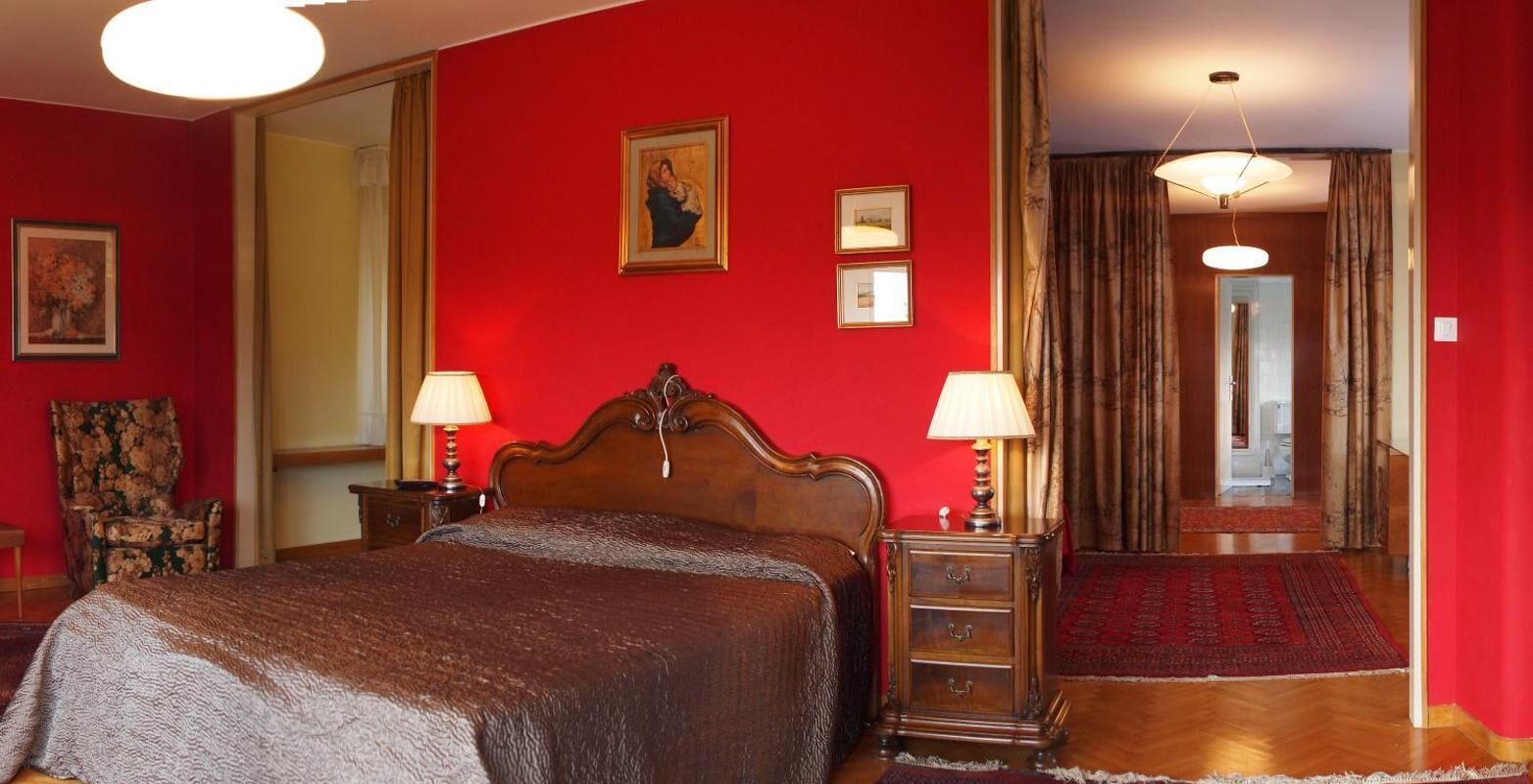 Quarto de hotel com uma cama grande, duas mesas de cabeceira com abajures, uma mesinha de madeira, parede vermelha e um espelho.