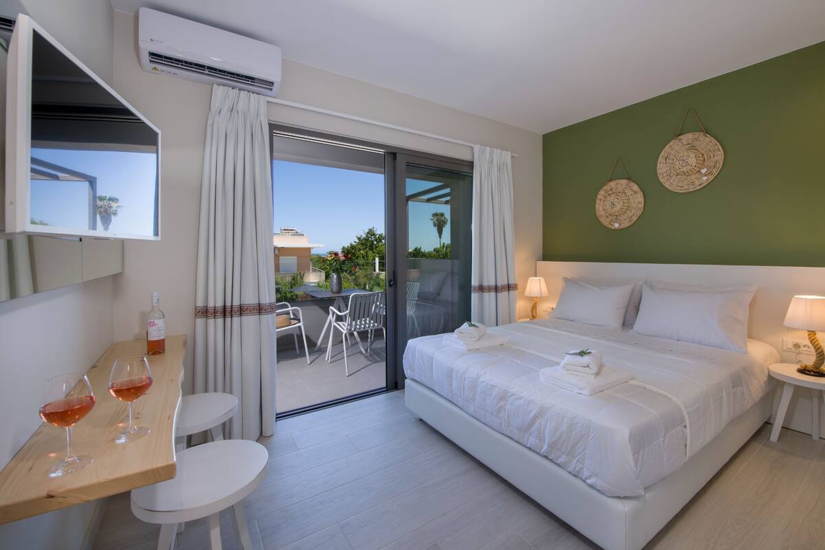 Quarto de hotel com uma cama, uma mesa com duas taças de vinho, uma cadeira e vista para o exterior.