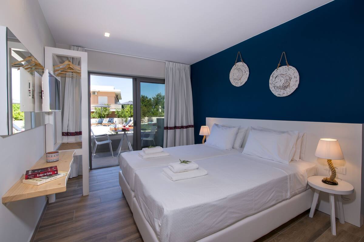 Quarto de hotel moderno com uma cama grande, uma mesa de cabeceira com abajur e vista para o exterior por uma porta de vidro deslizante.