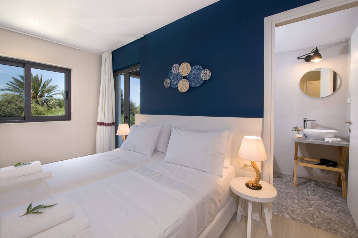 Quarto com uma cama grande, mesa de cabeceira com abajur, espelho redondo e vista para palmeiras através de uma janela.