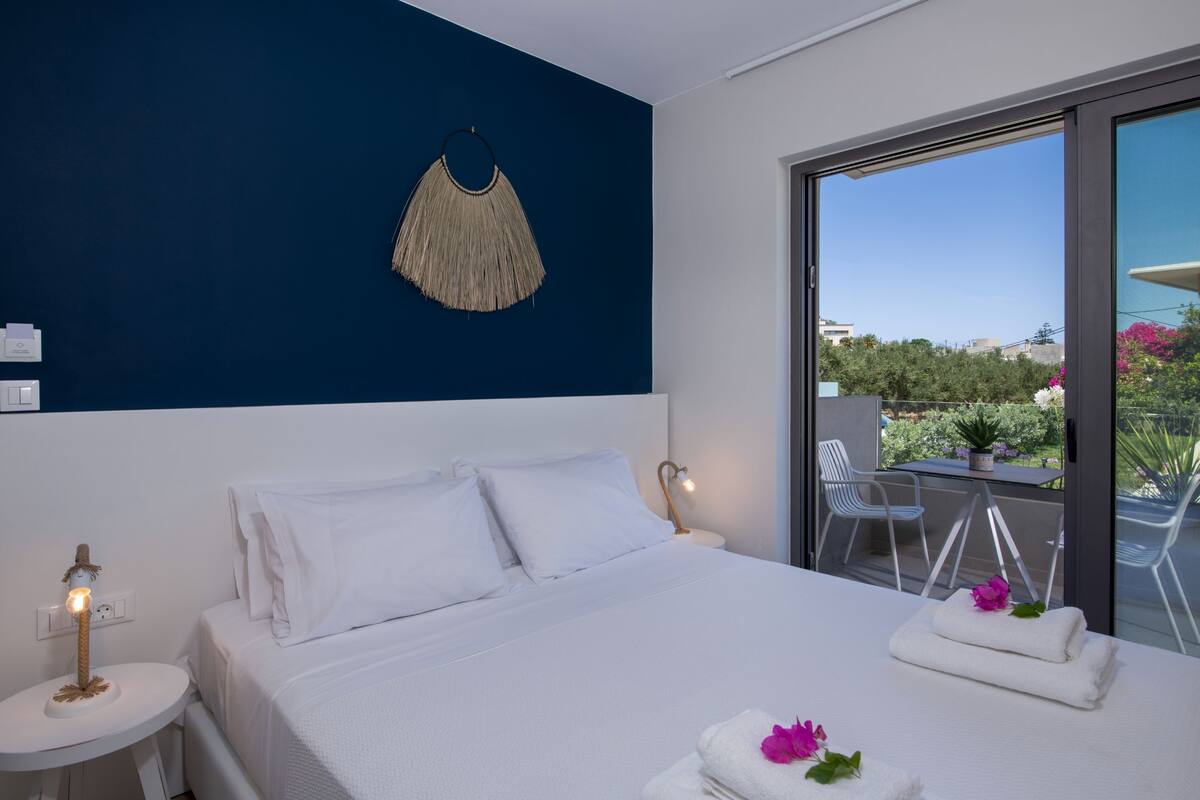 Quarto de hotel moderno com uma cama grande, uma pequena mesa de cabeceira com abajur, um painel decorativo e uma varanda com móveis de exterior.