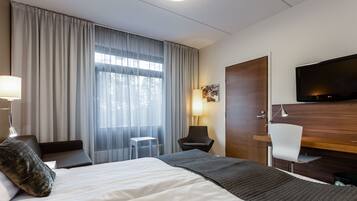 Quarto de hotel com uma cama grande, uma escrivaninha com cadeira, uma televisão e uma janela com cortinas.