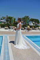 Uma noiva de vestido branco parada à beira de uma piscina, com cadeiras de descanso e guarda-sóis ao fundo.