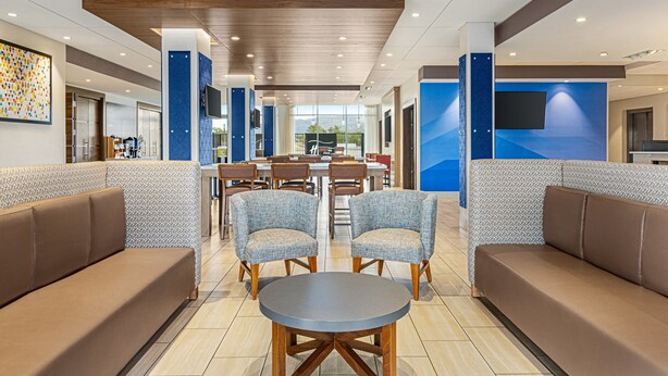 Um saguão moderno de hotel com área de estar central, uma mesa de jantar e uma tela grande.
