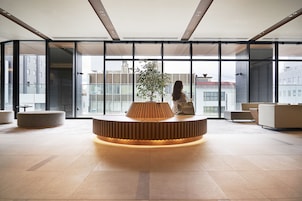 Um lobby moderno com uma mesa de recepção de madeira central, grandes janelas e assentos contemporâneos.
