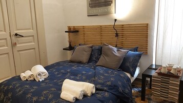 Cama bem arrumada com um edredom estampado floral, dois travesseiros e toalhas enroladas sobre um cobre-leito azul escuro.