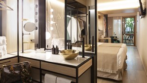 Quarto de hotel moderno com um espelho grande, toucador iluminado, pia dourada e uma cama com manta bege, além de vista para o exterior.