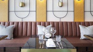 Uma sala de jantar com mesa posta para dois, apresentando um banco estofado marrom, uma mesa de mármore e luminárias pendentes fixadas na parede.