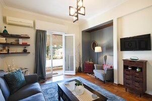 Sala de estar com sofá, mesa de centro e televisão. Há prateleiras com livros e objetos decorativos, um abajur e um armário vermelho.