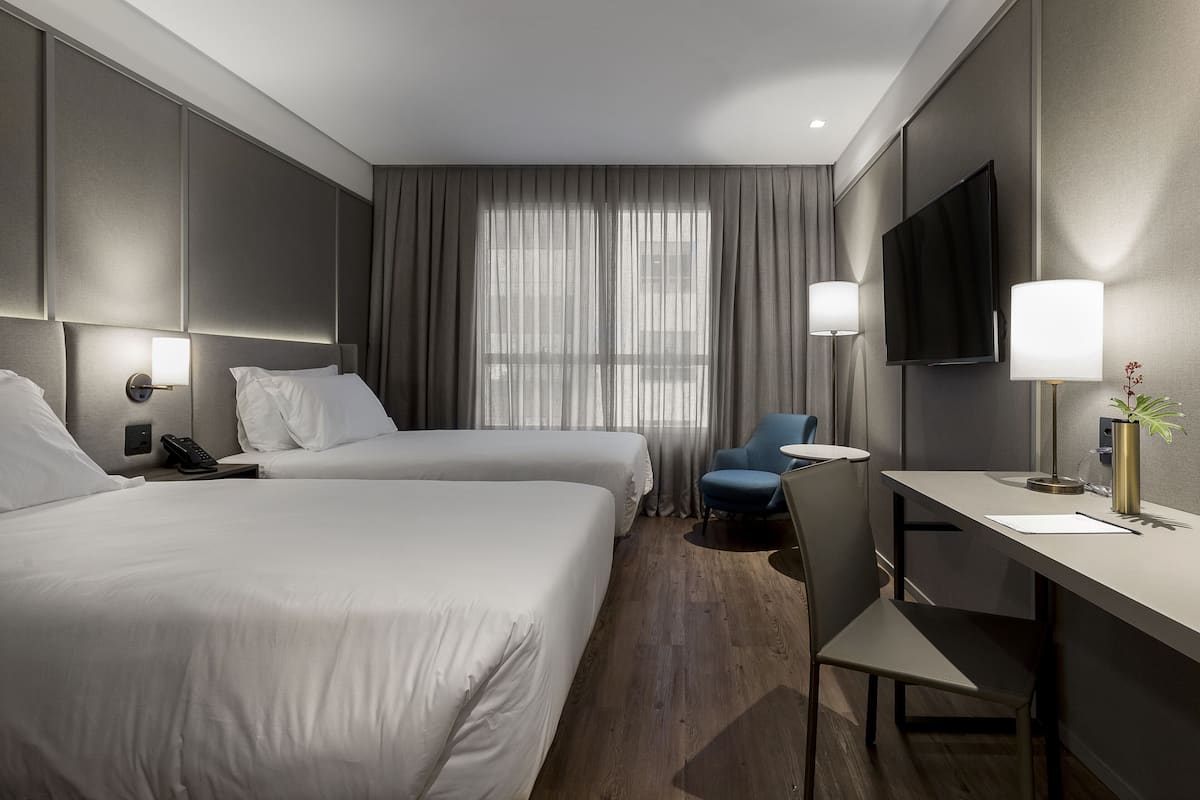 Quarto de hotel com duas camas, uma escrivaninha, uma cadeira, televisão e janela com cortinas.