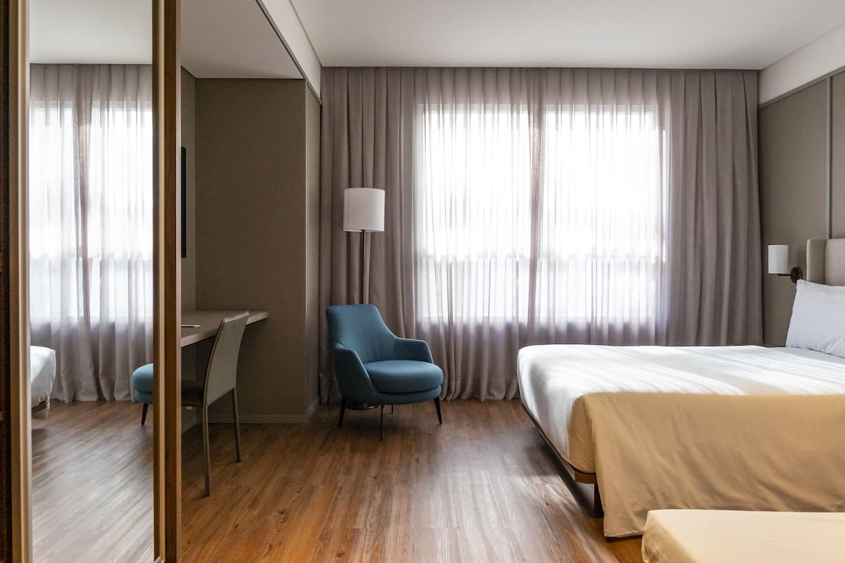 Quarto de hotel com cama, escrivaninha, cadeira e uma janela grande com cortinas.