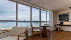 Quarto moderno com grandes janelas, uma mesa de madeira redonda e um espaço de estar aconchegante.