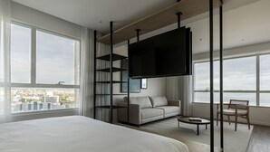 Quarto de hotel moderno com uma cama grande, um sofá, uma TV montada em uma prateleira de madeira e vista para a cidade através de amplas janelas.