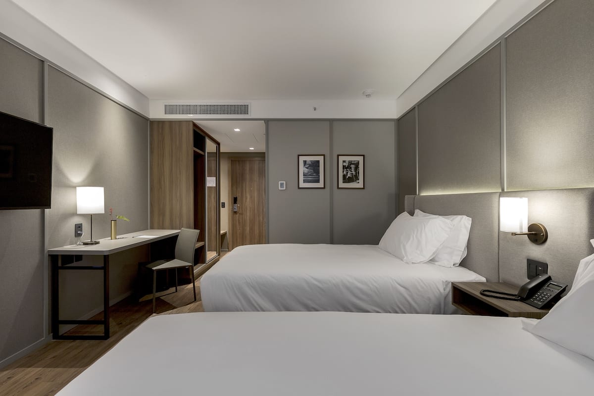 Quarto de hotel com duas camas, uma escrivaninha, uma cadeira e um telefone na mesinha de cabeceira.