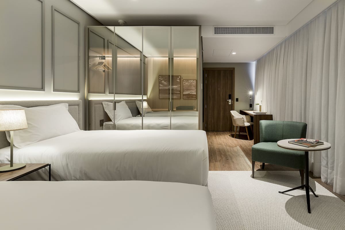Quarto de hotel com duas camas, uma poltrona verde, uma mesa pequena e um espelho.