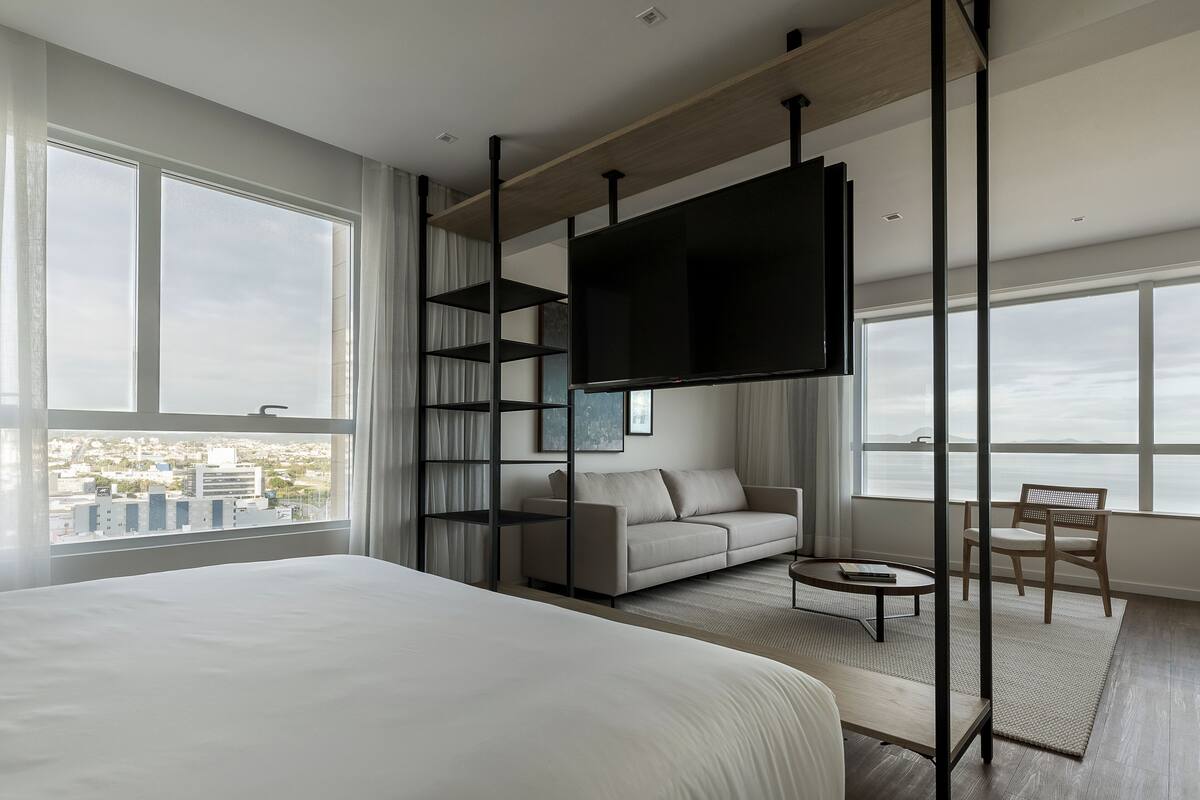 Quarto de hotel moderno com uma cama grande, um sofá, uma TV e vista para a cidade.