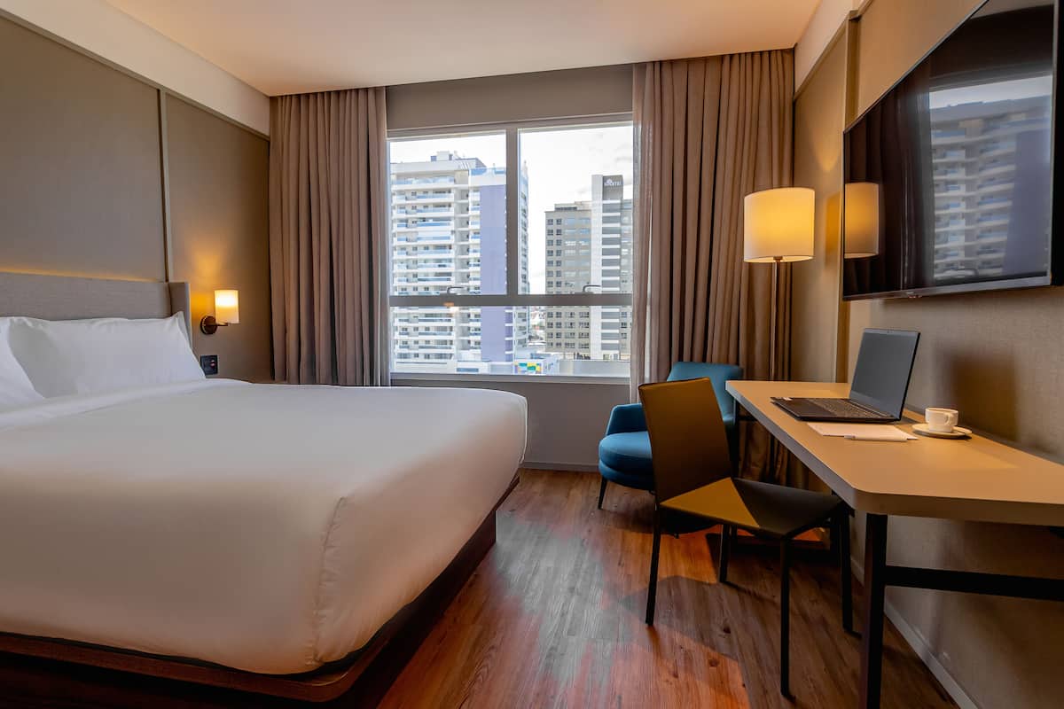 Quarto de hotel com uma cama grande, uma mesa com um laptop, uma cadeira e uma janela com cortinas.