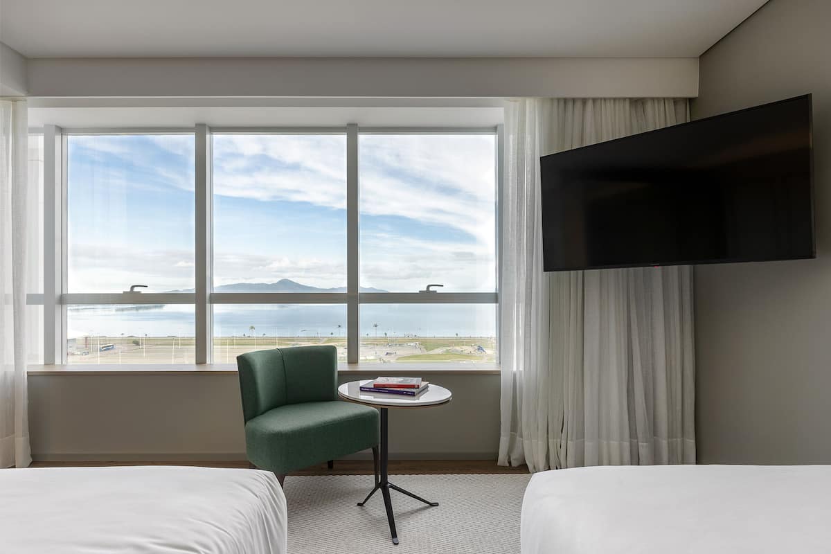 Quarto de hotel com duas camas, uma poltrona verde, uma mesa pequena com livros, uma TV fixada na parede e uma janela grande com vista para a praia e o oceano.