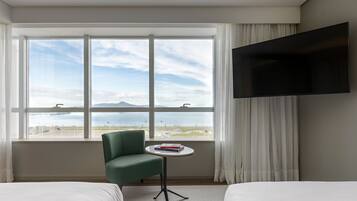 Quarto de hotel com duas camas, uma poltrona verde, uma mesa pequena com livros, uma TV fixada na parede e uma janela grande com vista para a praia e o oceano.
